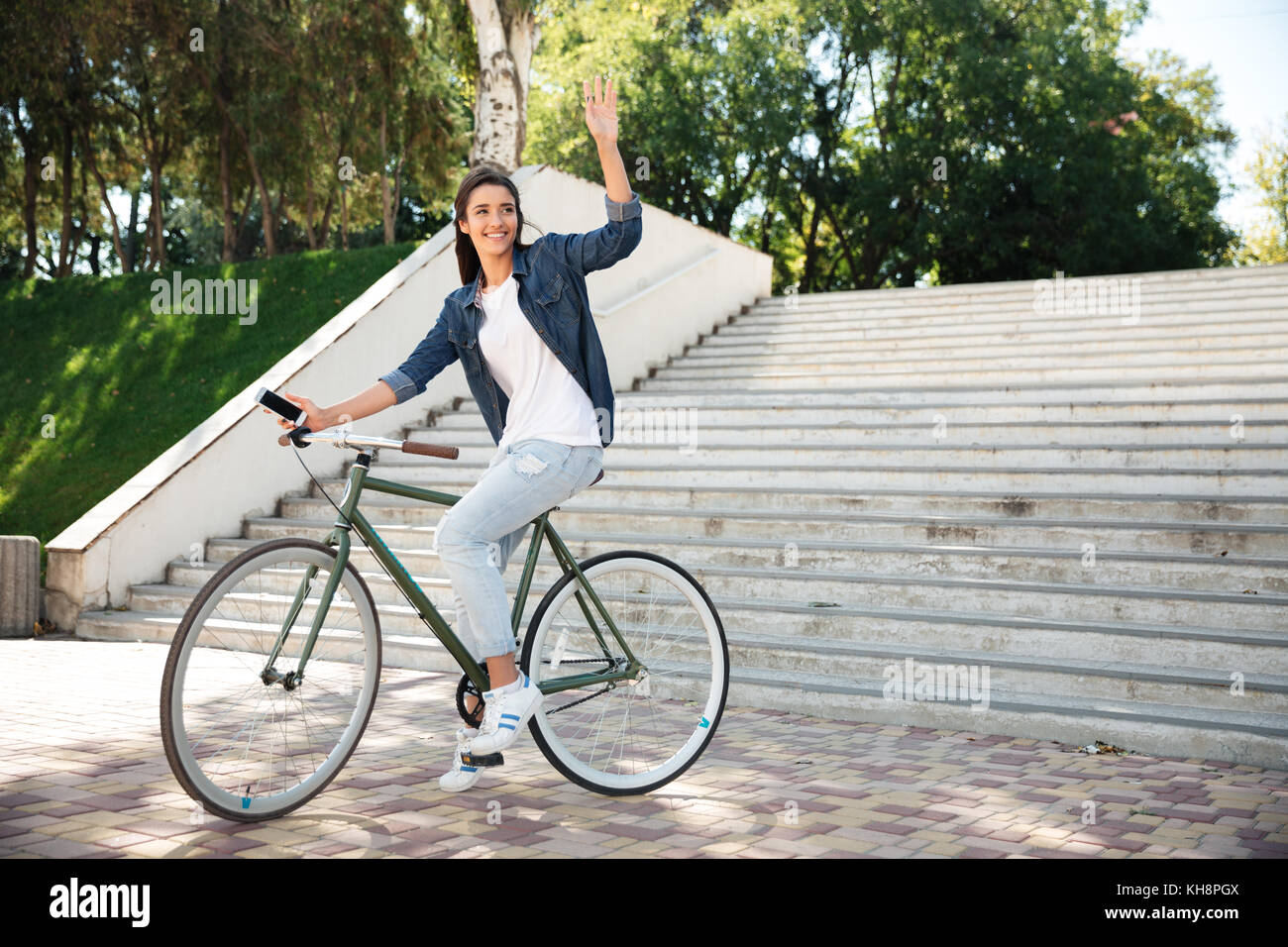 Portrait of a happy pretty girl waving hand while riding on a bicycle ...