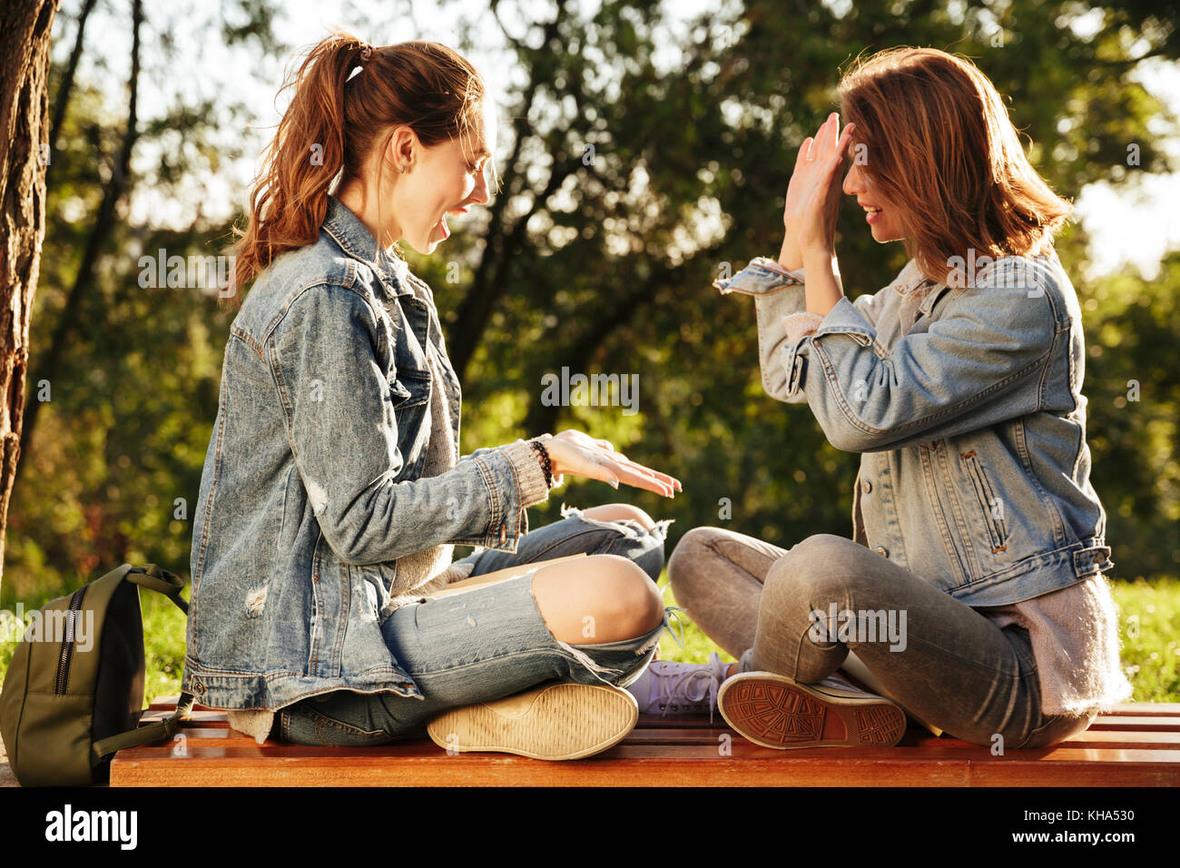 Two pretty girls clap their hands while sitting on wooden bench in park ...