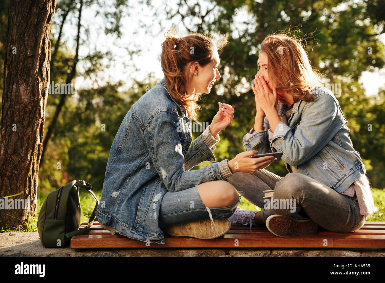 Two cheerful female friends sitting on wooden bench with crossed legs ...