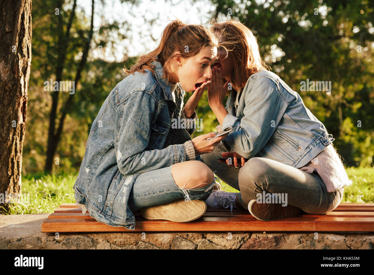 Two young pretty woman whispering secrets while sitting on wooden bench ...