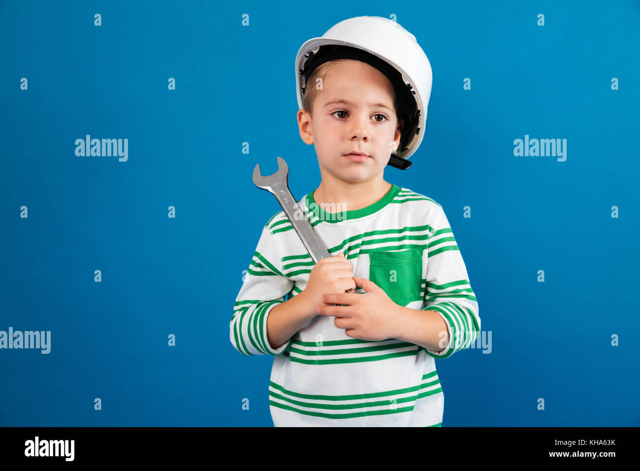 Bored young boy in protective helmet posing with wrench like engineer ...