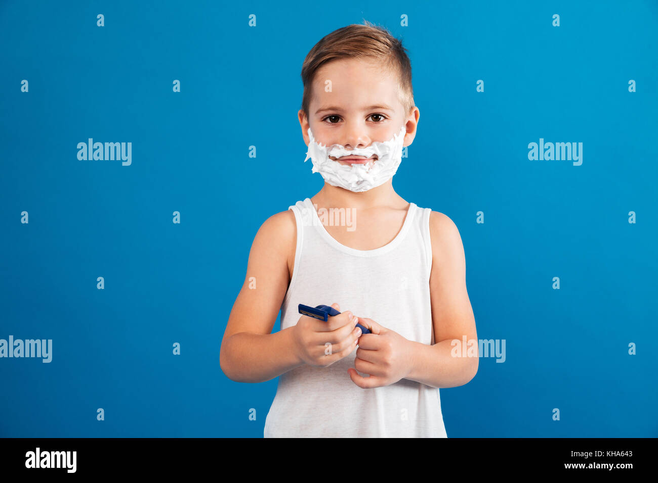 Smiling young boy in shaving foam holding razor like man and looking at ...