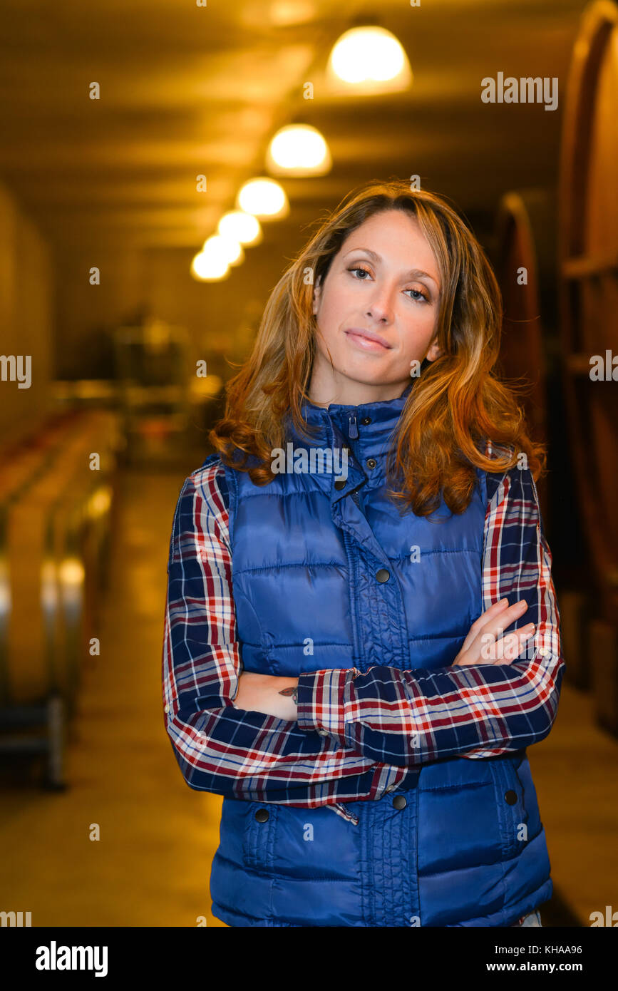 cheerful young woman winery owner posing in her cellar with wine barrel ...