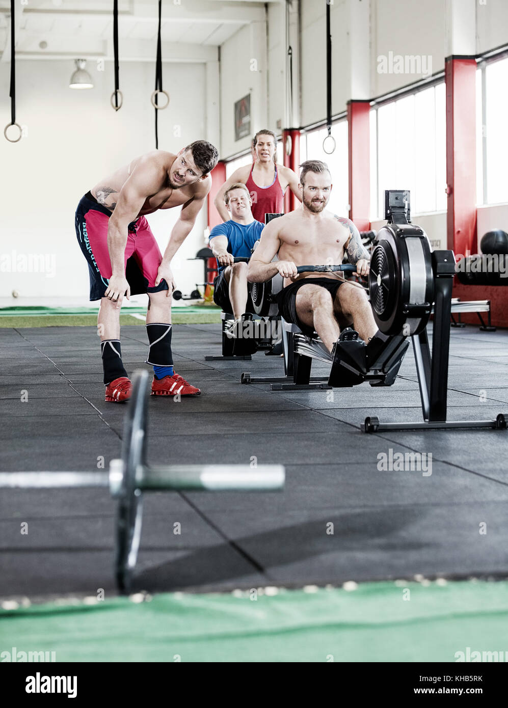 Tired young man standing by friends exercising on rowing machine in gym ...