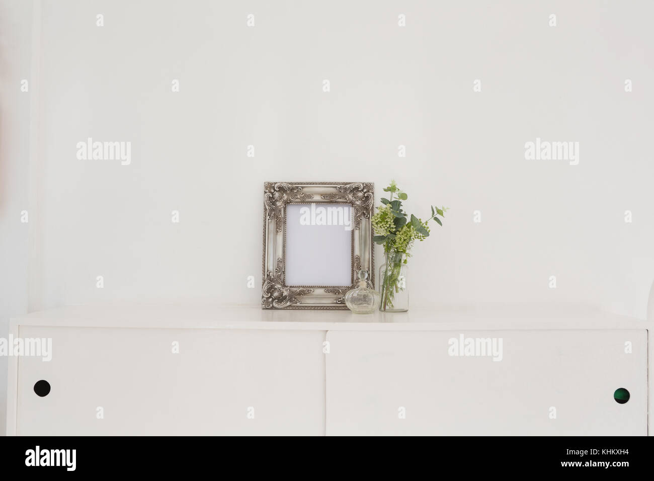 Picture frame, perfume bottle and flora on table against white wall ...