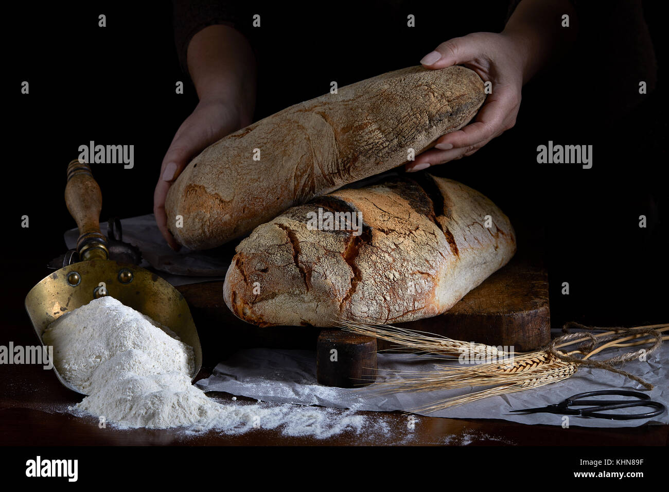 Bread loaf in baker hands on rustic table with flour and wheat ears, dark moody setting Stock ...