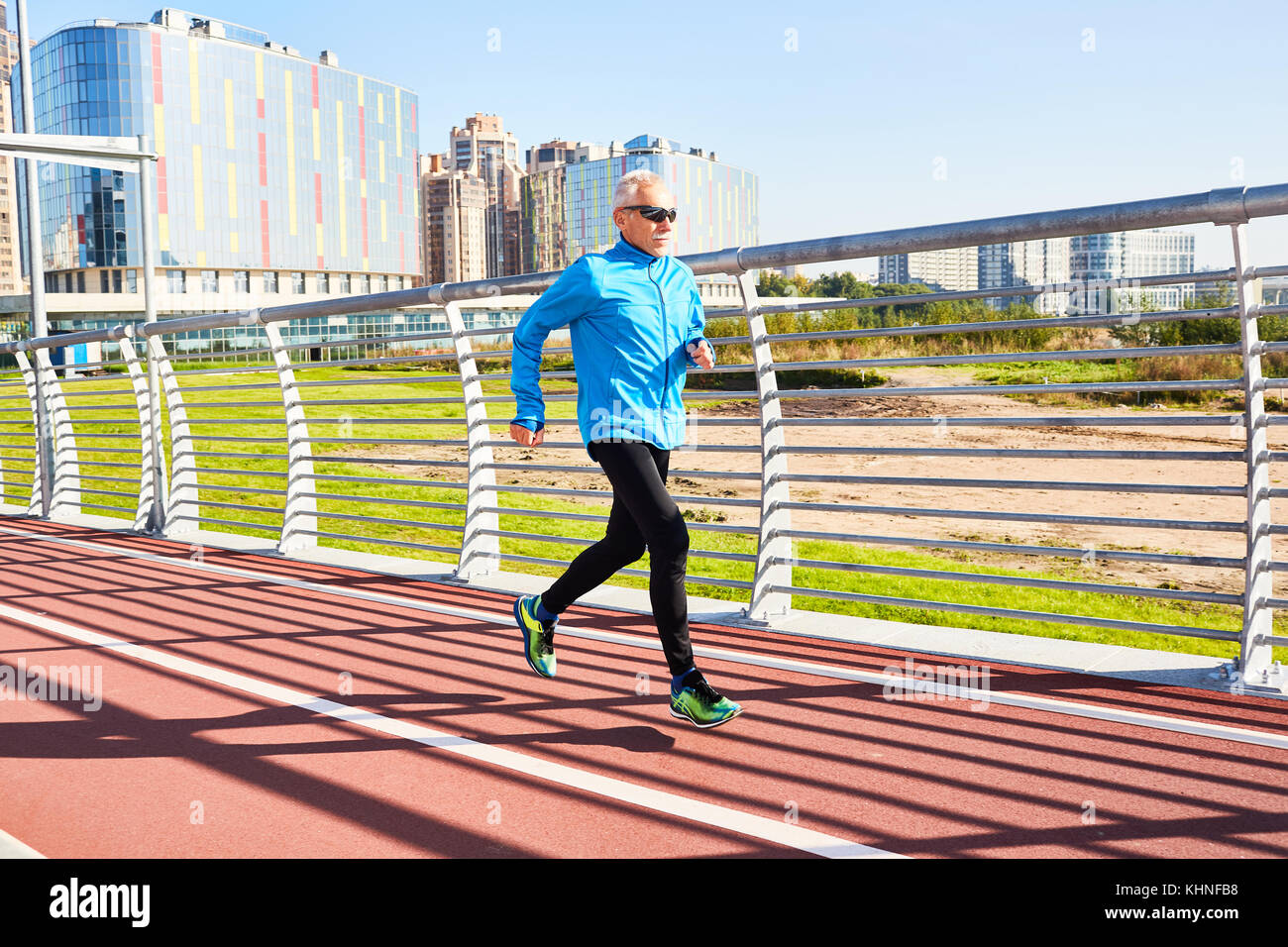 Retired man in activewear jogging along railings with cityscape behind ...