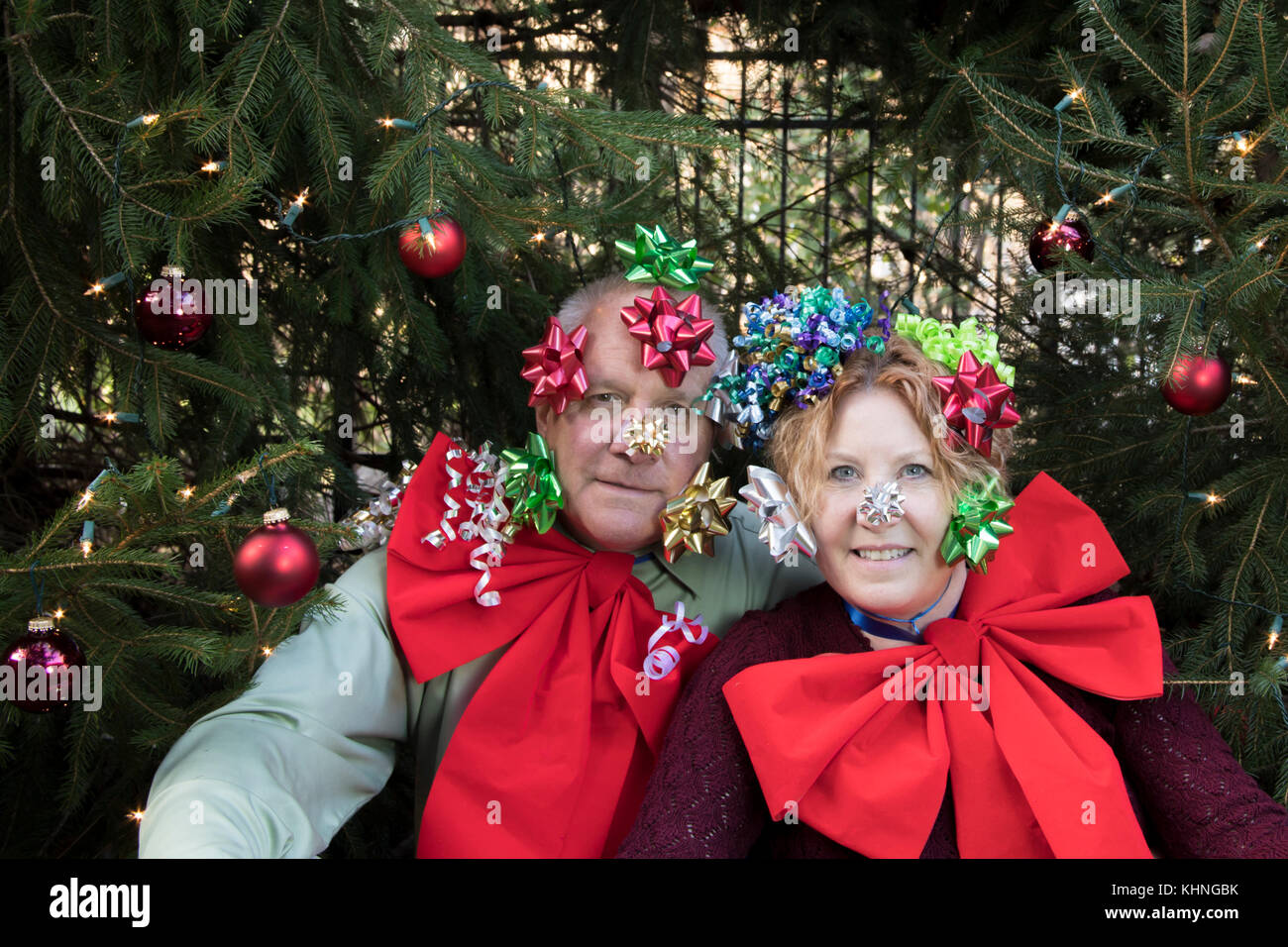Couple covered in Christmas bows sitting under pine trees decorated ...