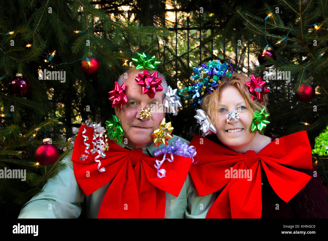 Couple covered in Christmas bows sitting under pine trees decorated ...