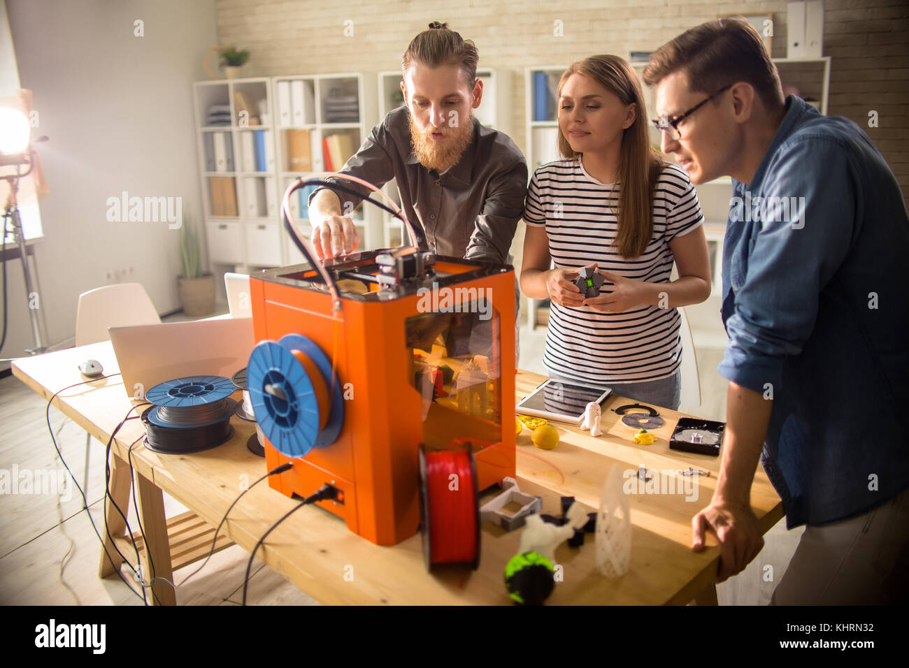 Three young engineers standing at wooden desk and using 3D printer in ...