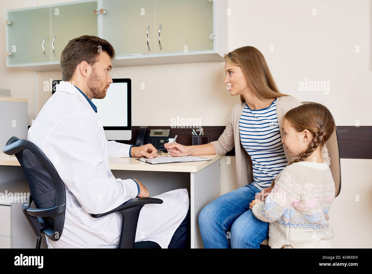 Portrait of little girl visiting doctor with mother, sitting at desk in ...