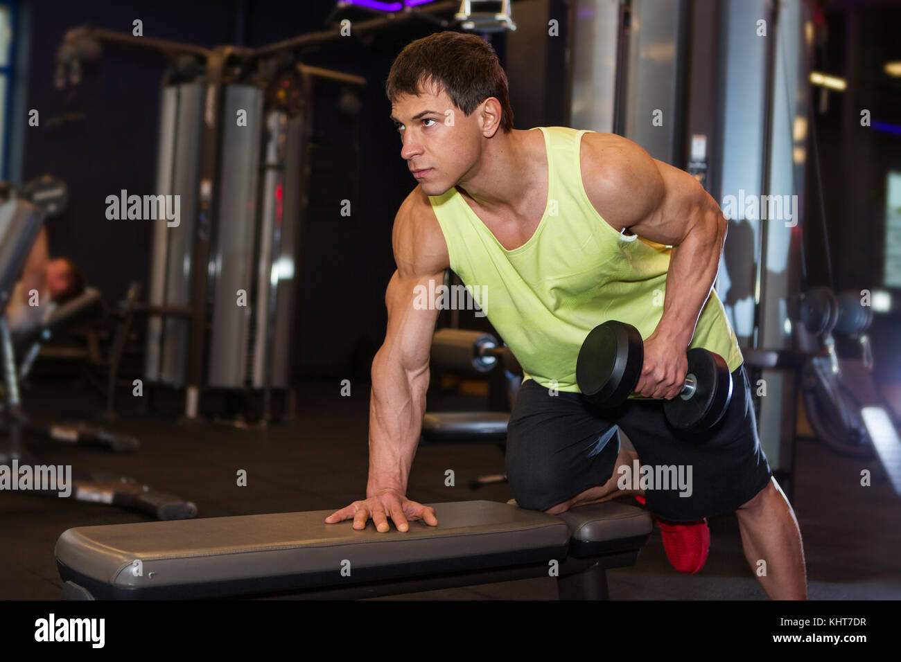 Young muscular man in a vest doing one-arm dumbbell rows on bench in ...