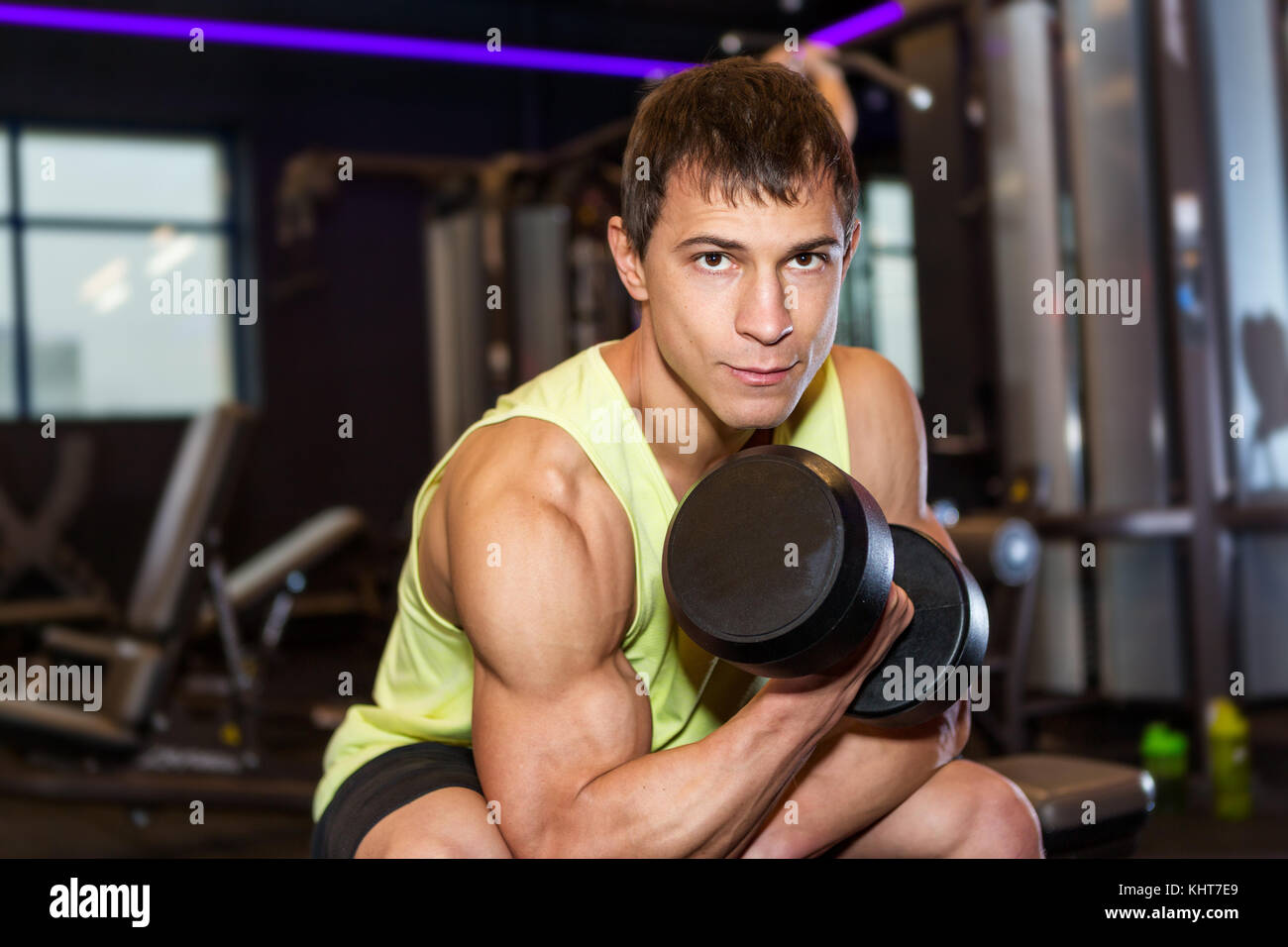 Young muscular man in a vest doing one-arm dumbbell rows on bench in ...