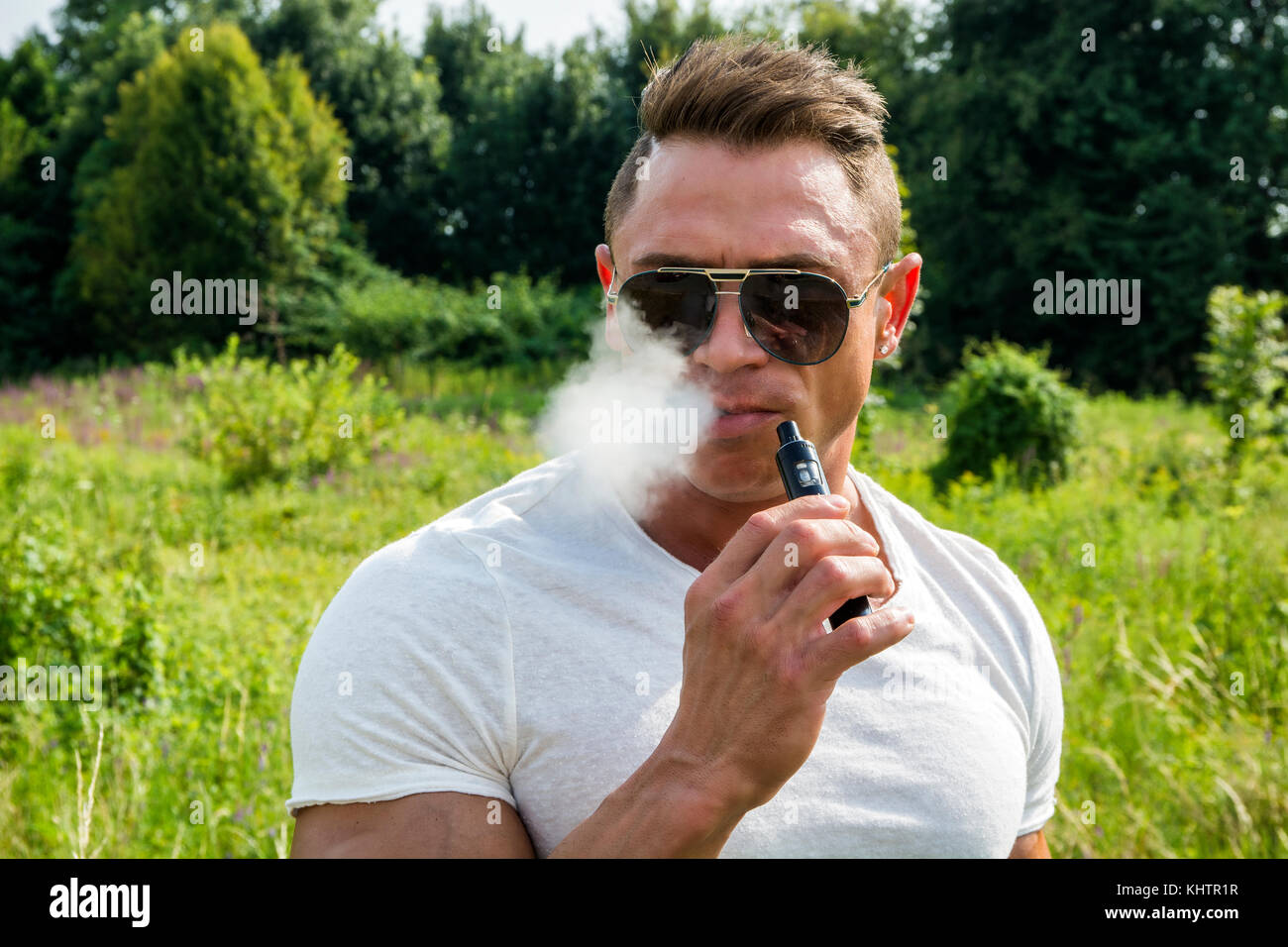 Handsome man standing in nature and vaping with electronic cigarette ...