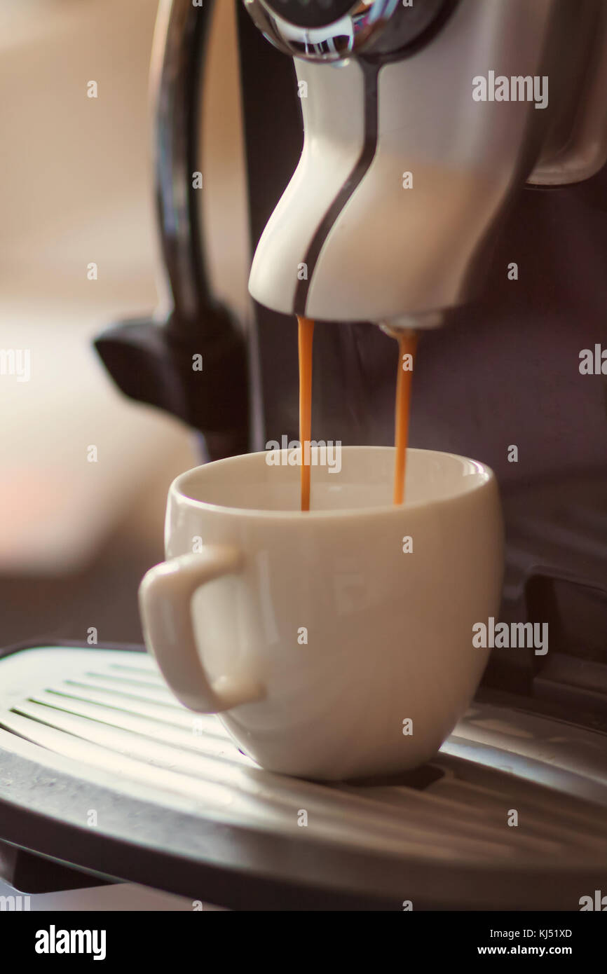 White cup standing on the grating of coffee machine with coffee pouring ...