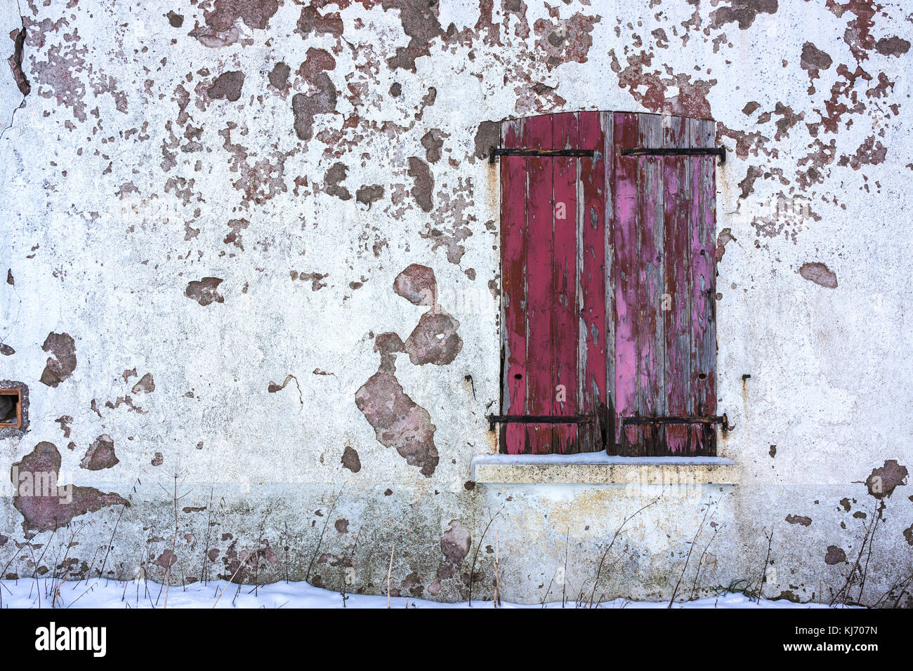 An old window with closed red worn wooden shutters on a white decrepit ...