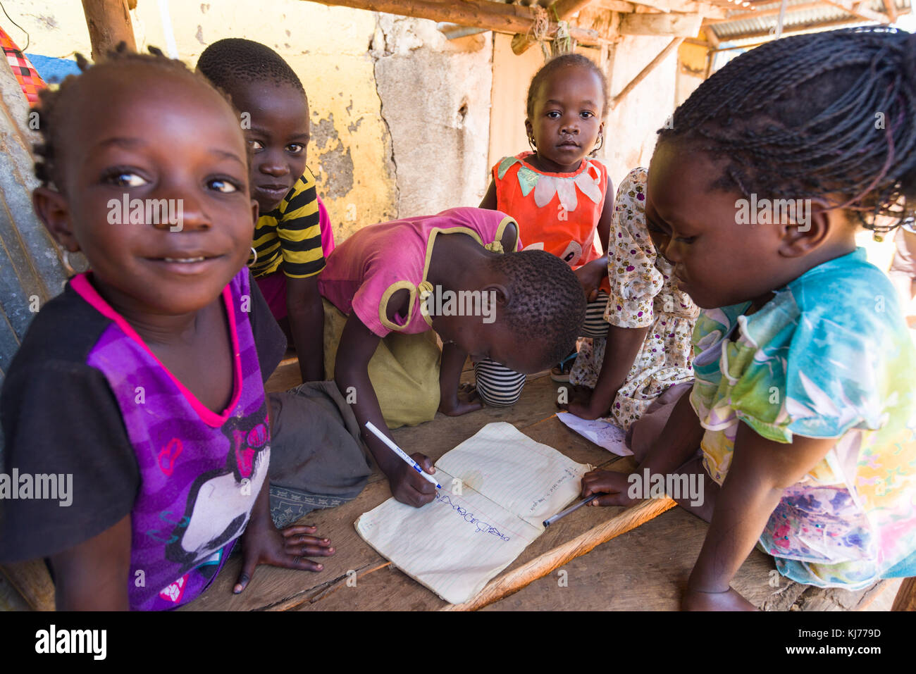 A group of young children sit around an exercise book in the shade of a ...