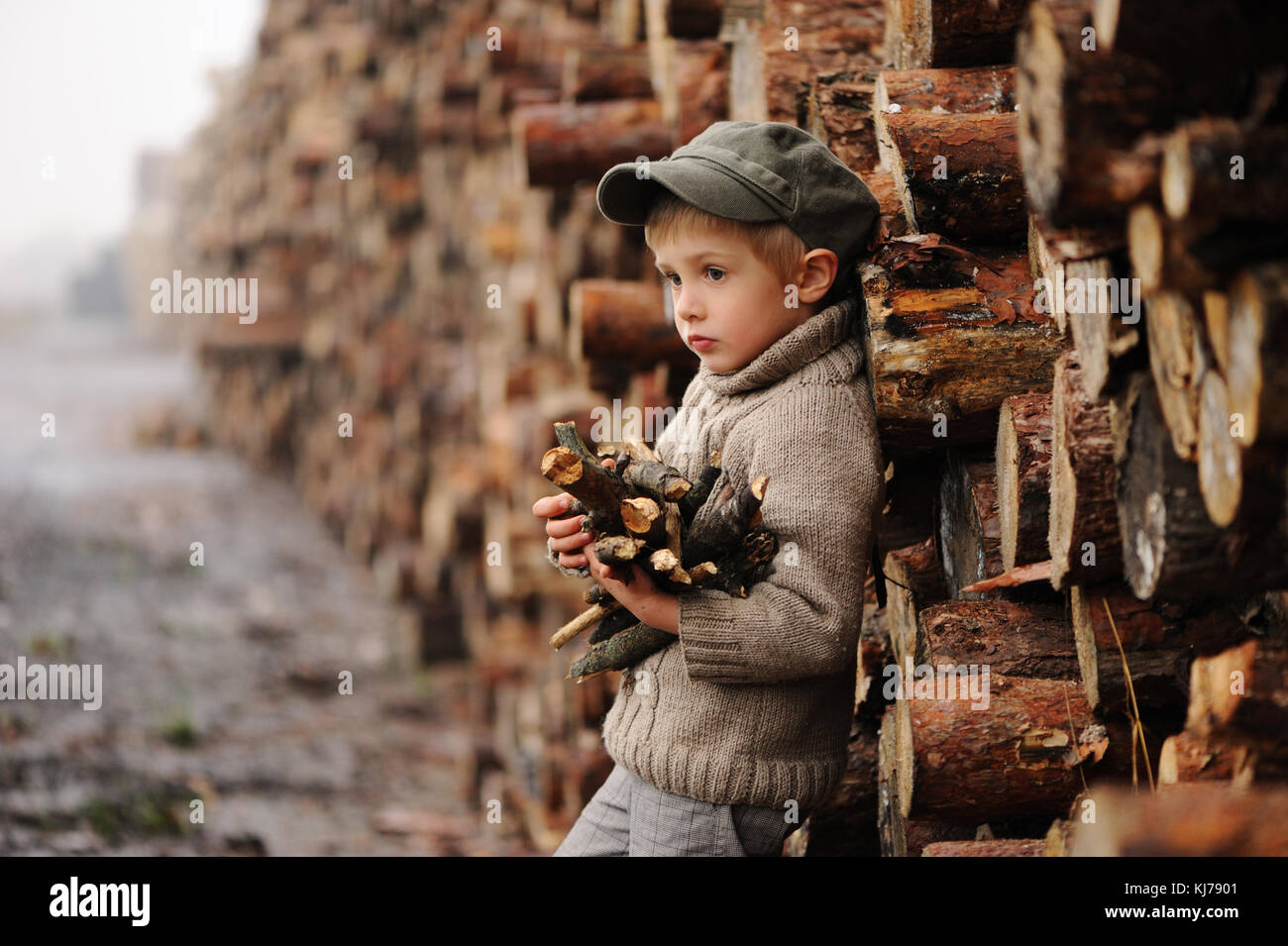 A sad,pretty, four years old boy lumberjack playing on pill of wood ...