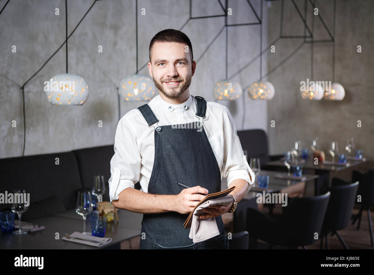 A young, smiling waiter in a restaurant, standing next to the tables ...