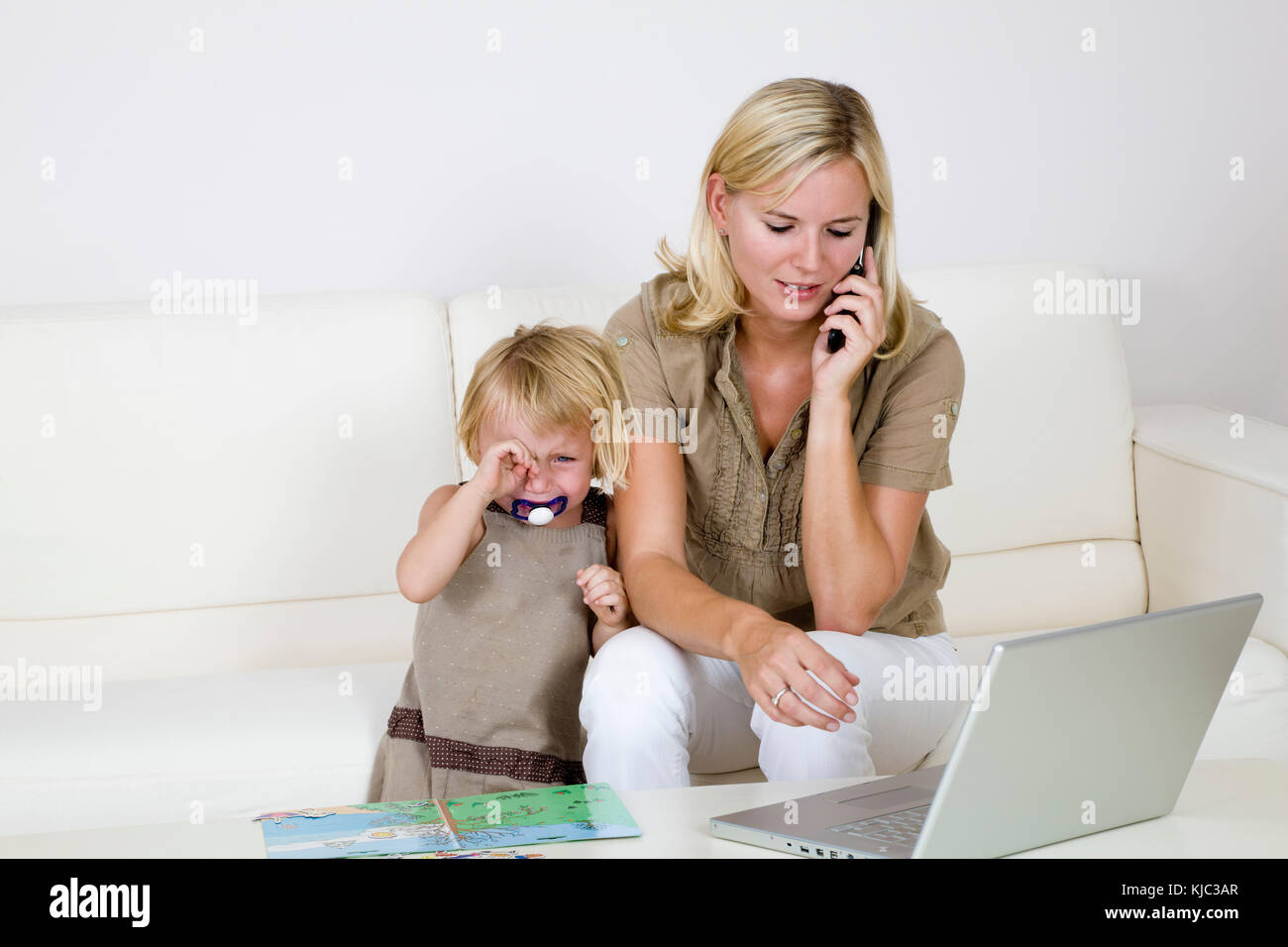Mother Using Laptop and Cell Phone, Toddler Standing Beside Her Crying ...