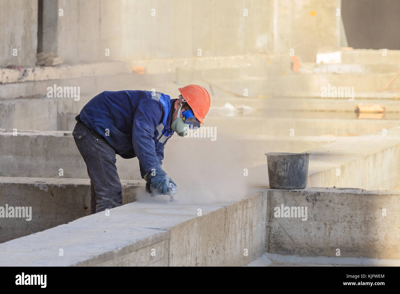 The worker in overalls and a protective mask grinds the concrete wall ...
