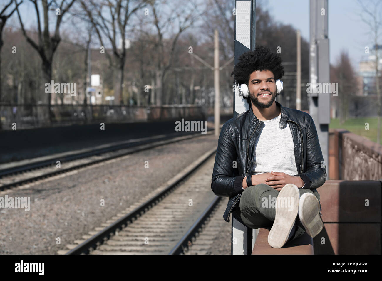 Smiling man sitting on wall listening to music next to train rails ...