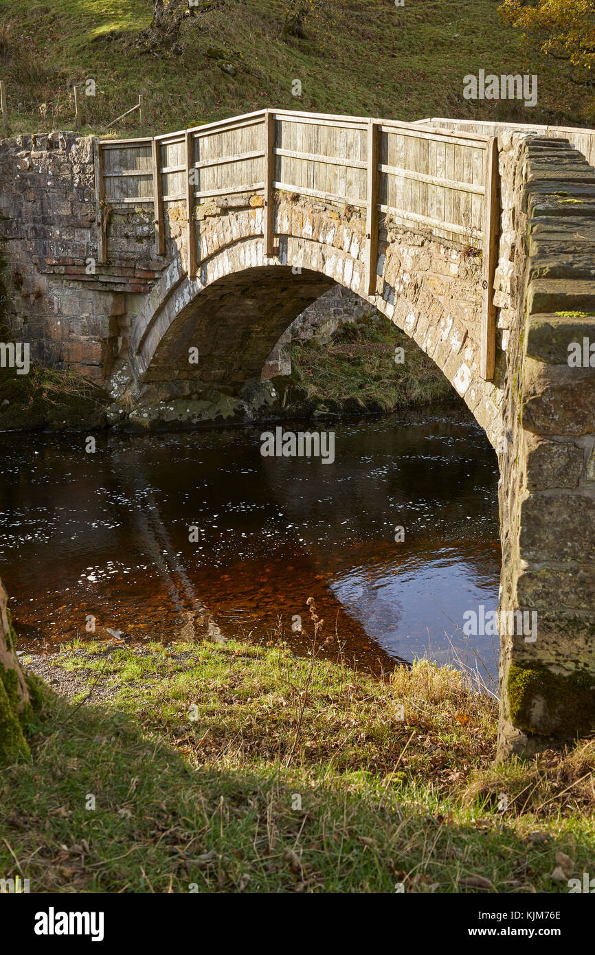 In afternoon autumn sunshine a stone bridge with wooden parapets spans ...