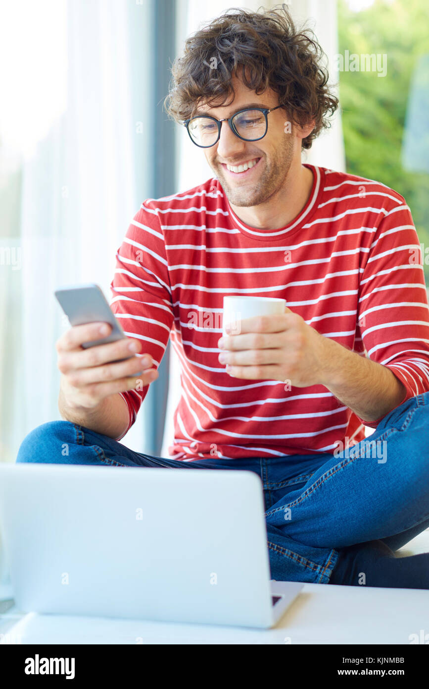Full length shot of a young man reading messages and drinking morning ...