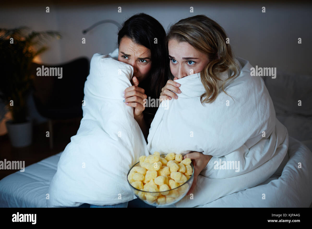 Scared girls hiding in blanket while watching horrors on TV at night ...
