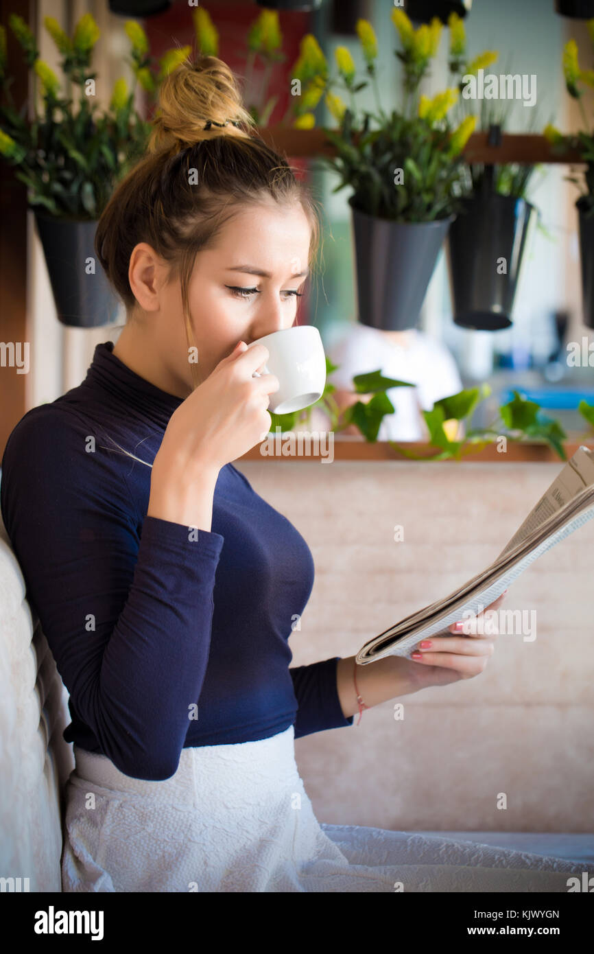 Beautiful young woman reading newspaper and drinking coffee while using ...