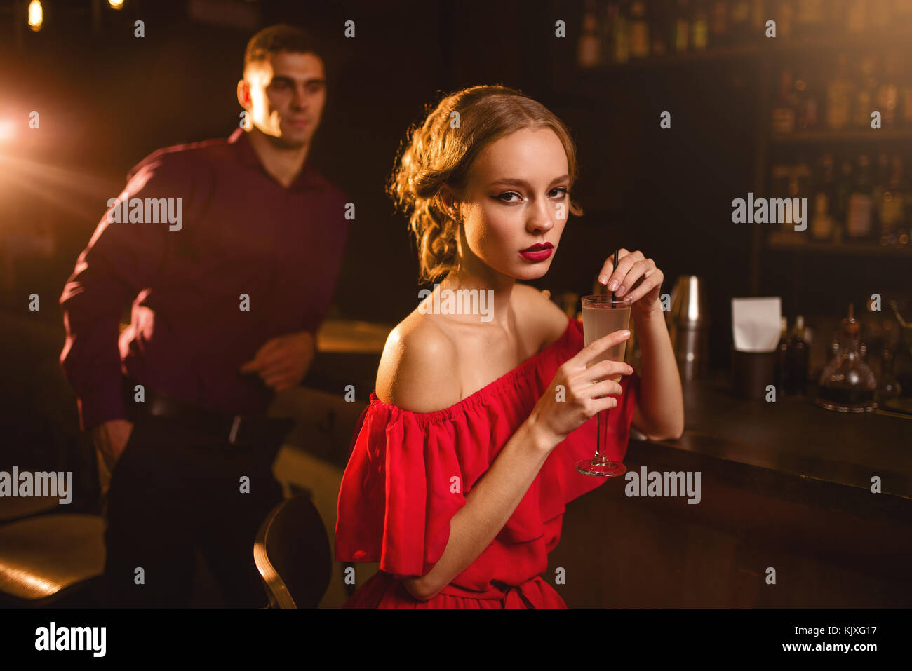 Woman in red dress with cocktail in hand, man behind bar counter, flirting. Date in nightclub ...
