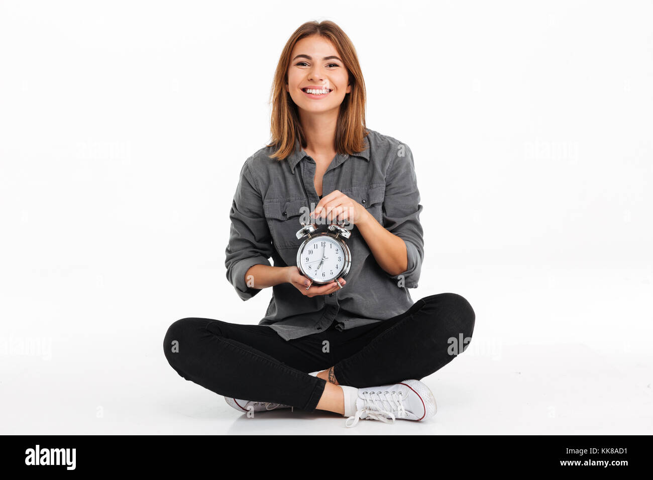 Portrait of a smiling pretty girl showing alarm clock while sitting and ...
