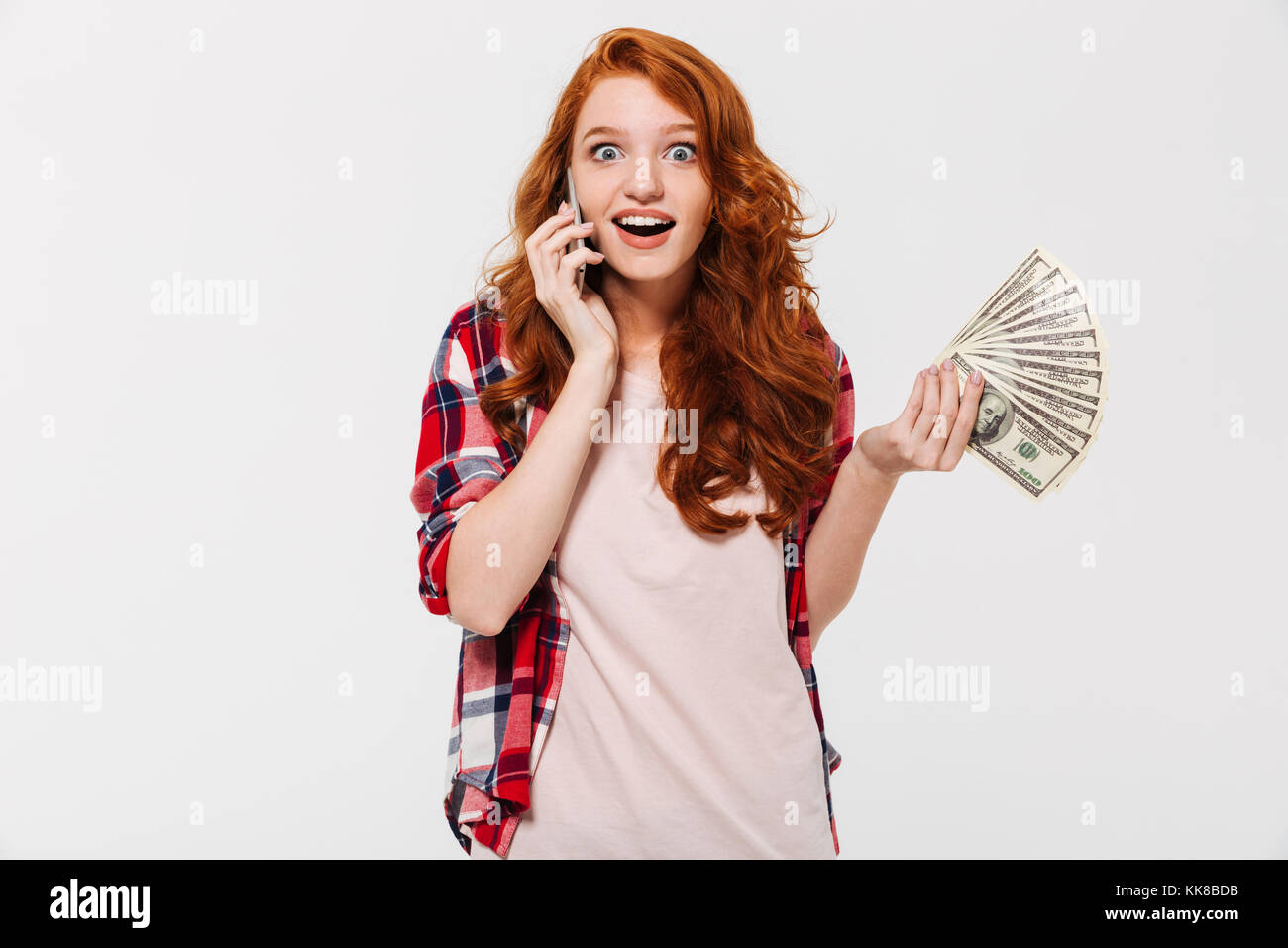 Happy Shocked ginger woman in shirt talking by smartphone while holding ...
