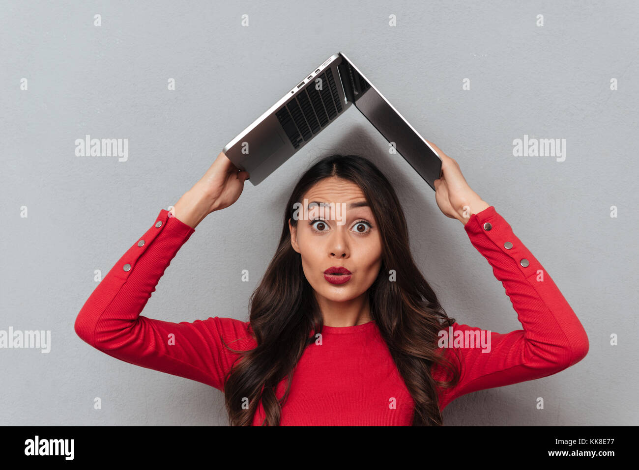 Shocked brunette woman in red blouse holding laptop computer overhead ...
