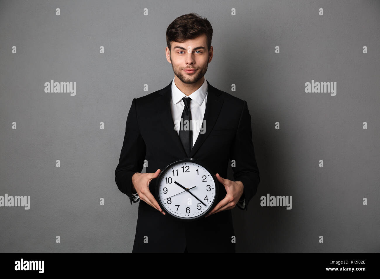 Young cheerful businessman in black suit holding big clock, looking at ...