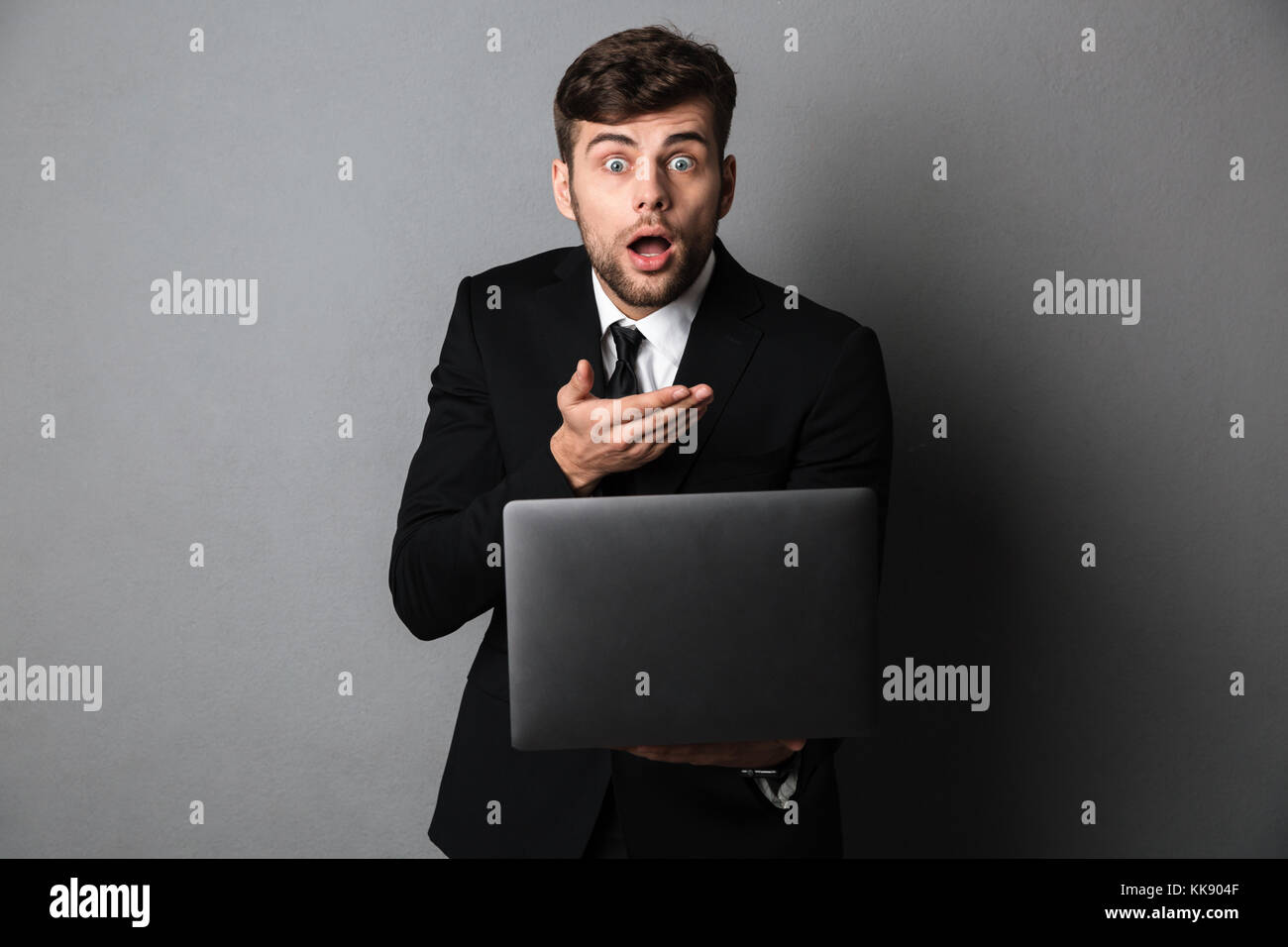 Amazed young guy in black suit holding laptop, looking at camera ...