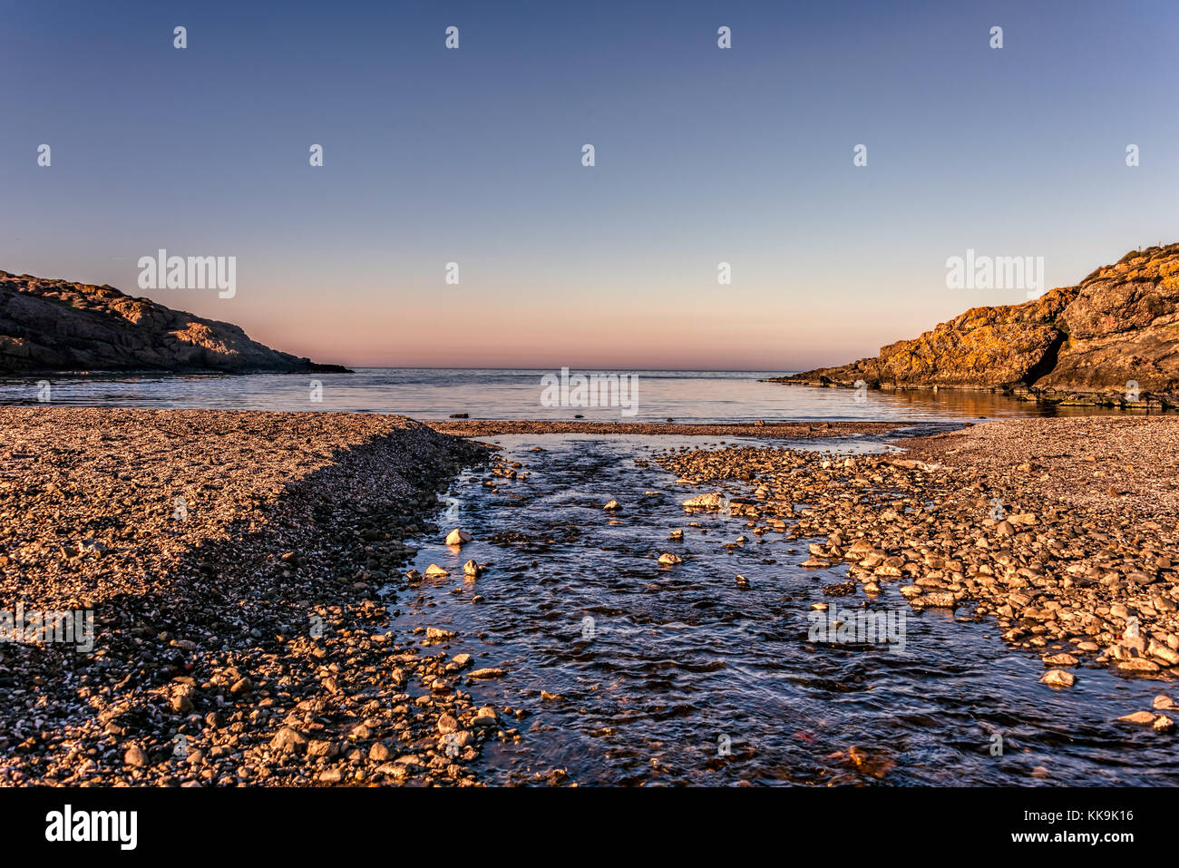 Pebble beach with a stream of water flowing into a still sea under blue ...