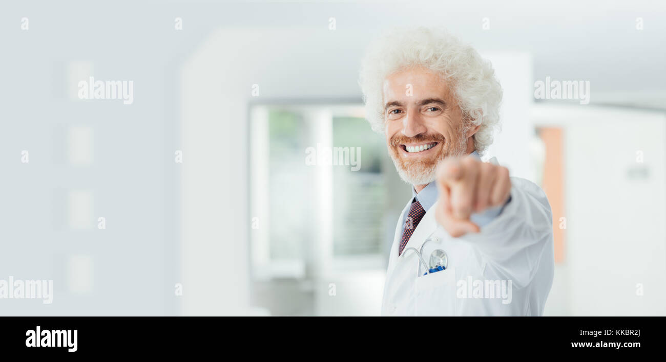 Smiling cheerful doctor pointing at camera, hospital interior on ...