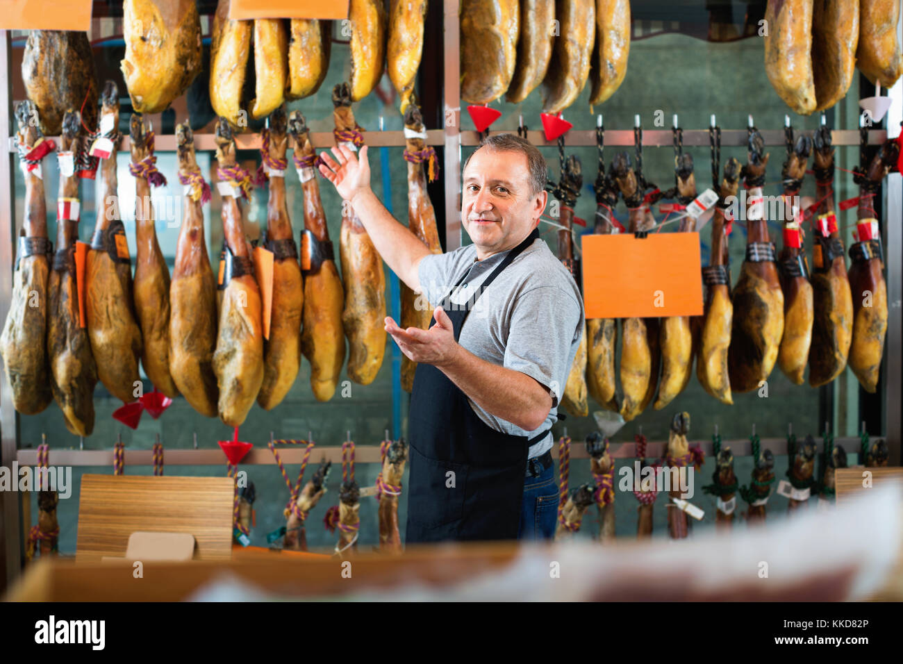 Portrait of cheerful mature shop man offering Spanish jamon in deli ...