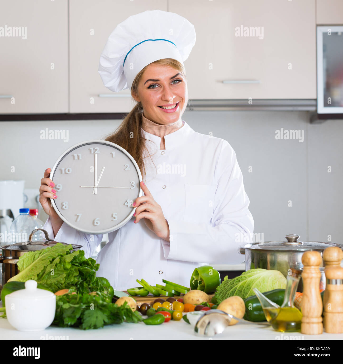 Smiling woman in chef cap holding clock as preparing vegetables Stock ...