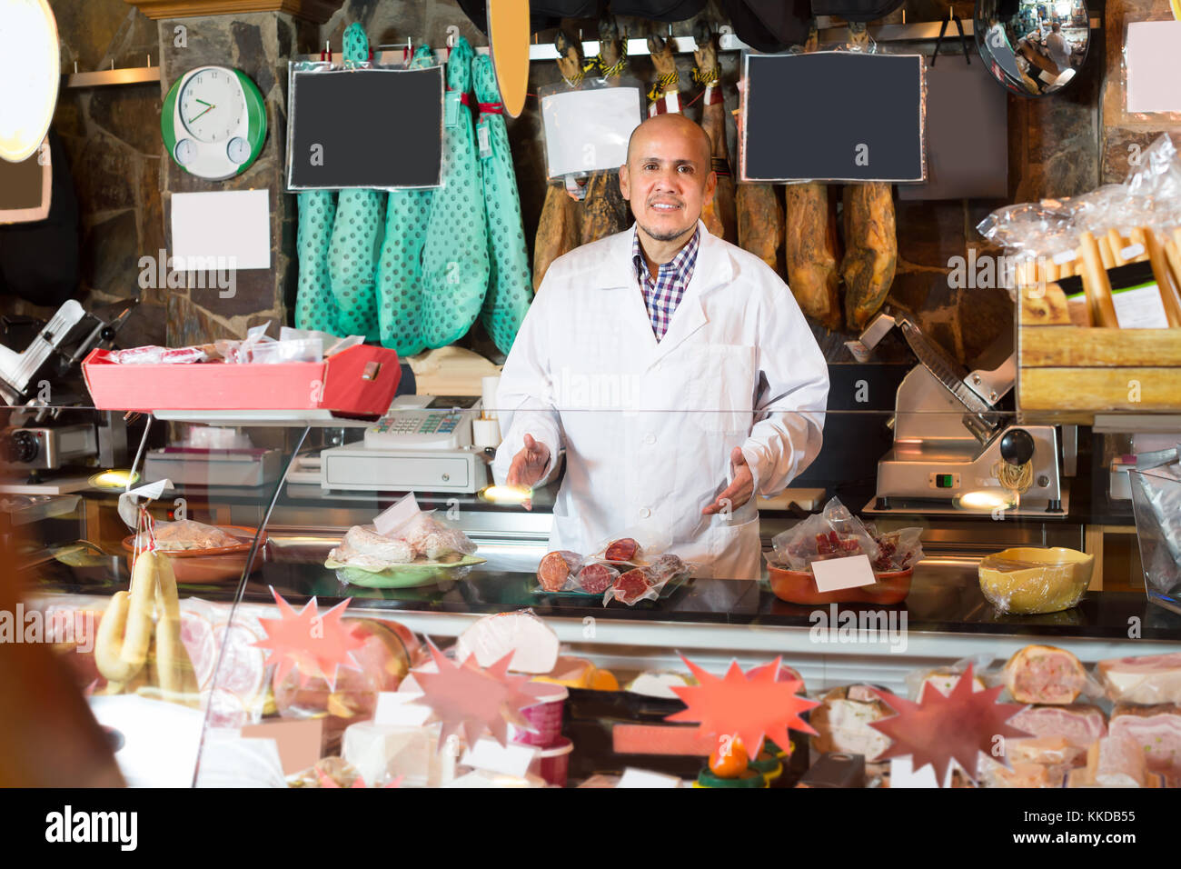 Portrait of positive male seller in white overall offering jamon and ...
