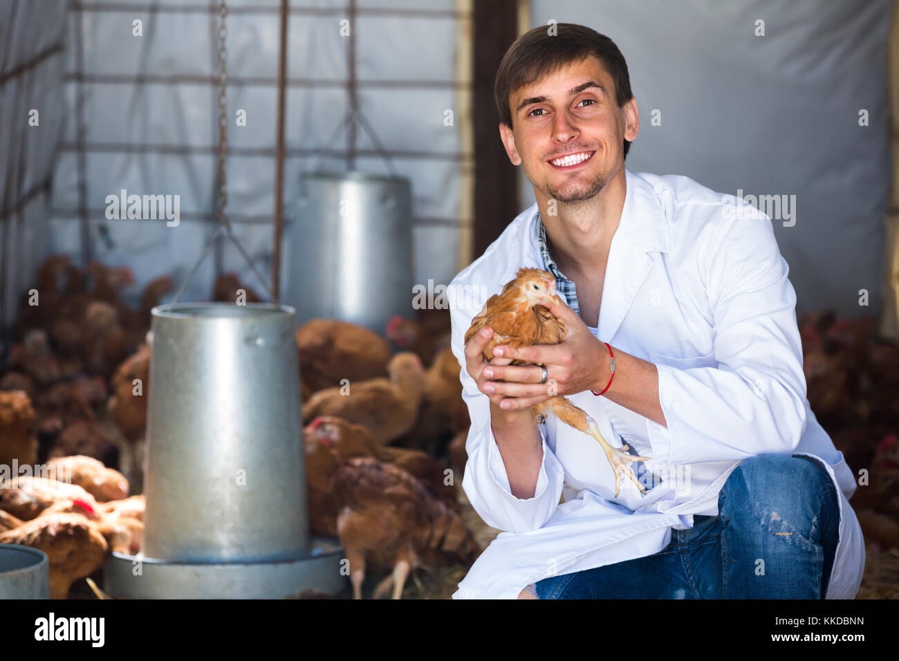 Joyful smiling young man in white coat having chicken in hands in hen ...