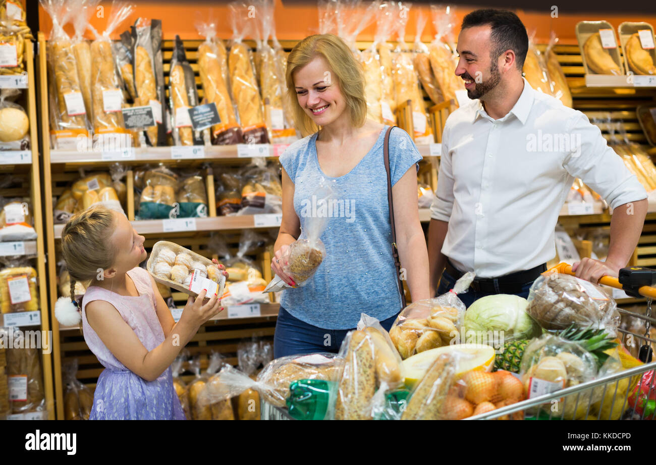Joyful young parents with little daughter picking bread from shelf in ...