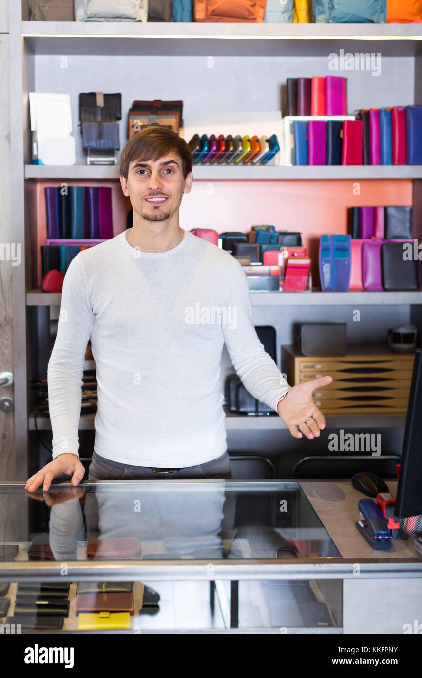 Smiling male shop assistant posing near glass display in wallet section ...