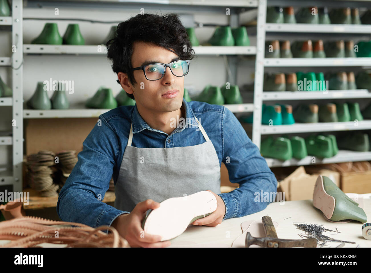 Young shoemaker in apron and eyeglasses sitting by workplace on ...