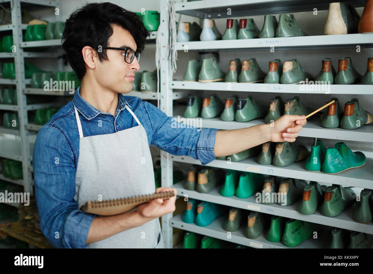 Young modern shoemaker pointing at pair of footwear workpieces on shelf ...