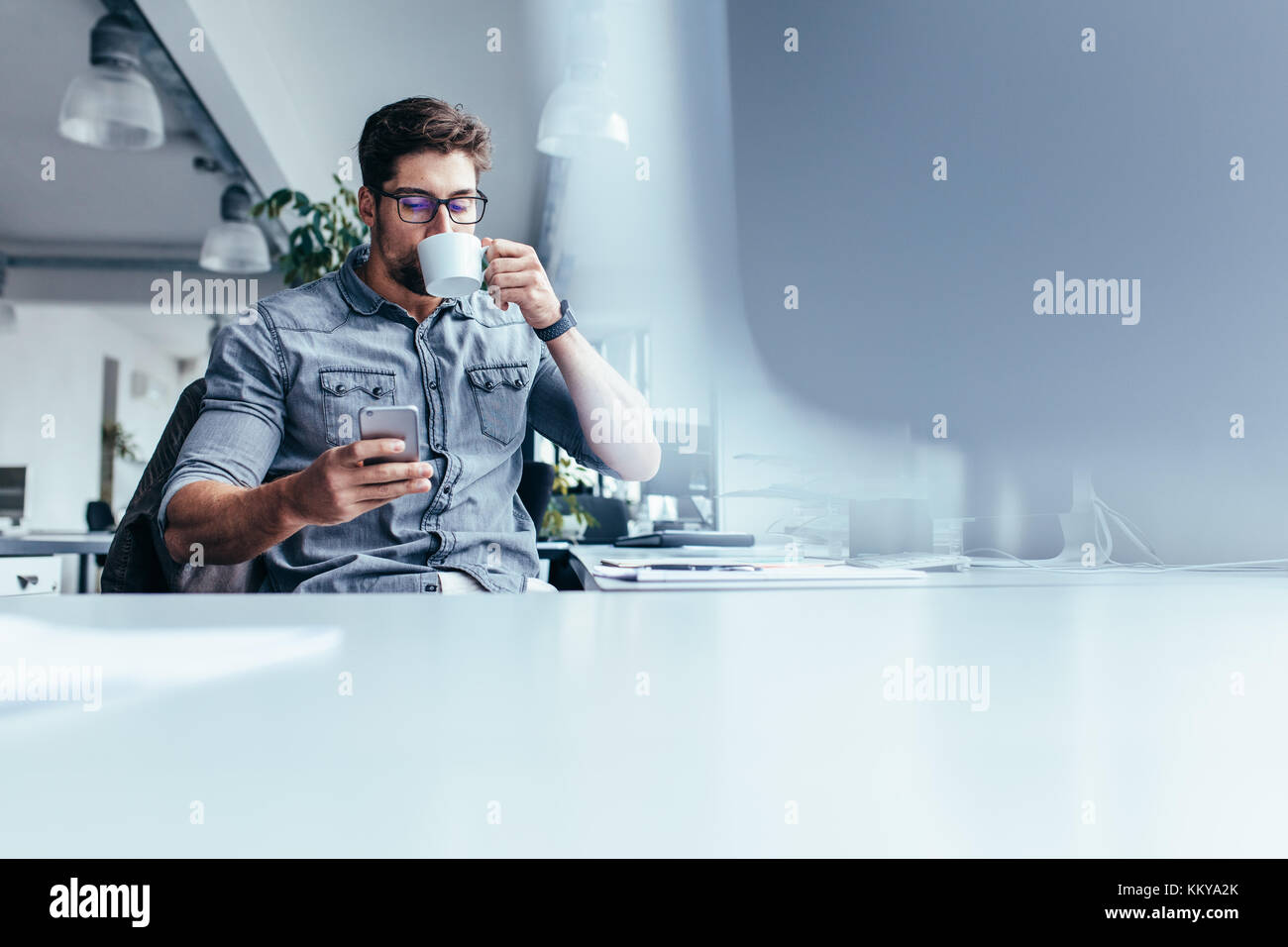 Male designer drinking coffee while reading text message on smart phone ...