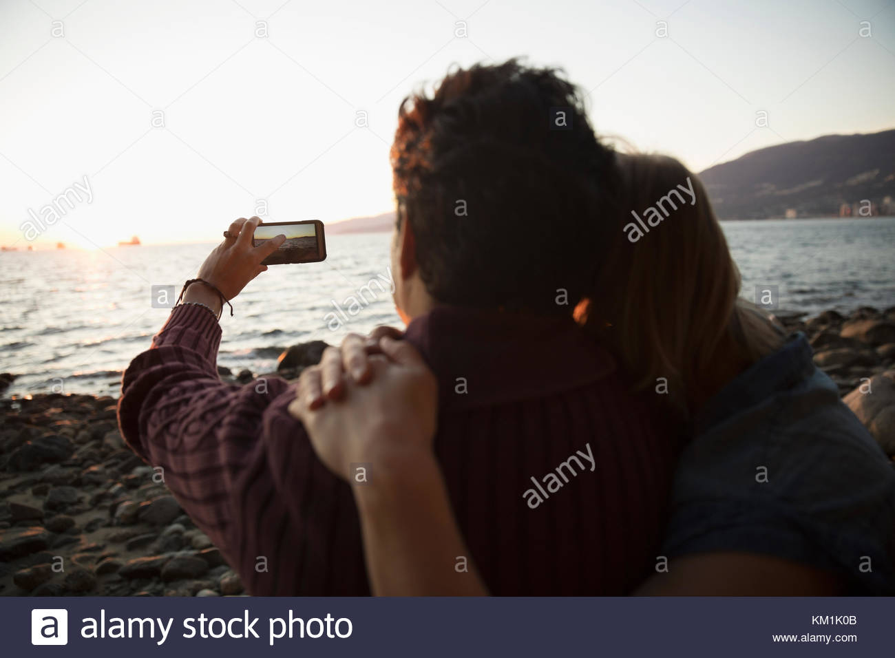 Man Photographing His Wife On Beach With Camera Phone Hi Res Stock
