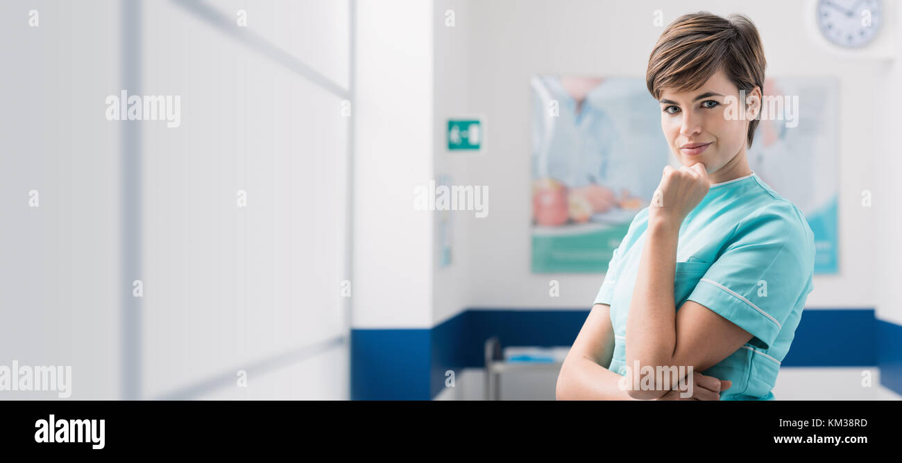 Confident young female nurse posing in the hospital aisle and smiling ...