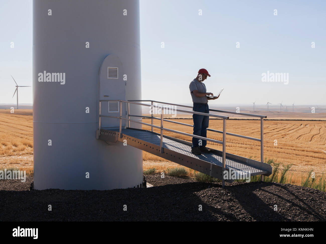 A wind farm technician standing and using a laptop at the base of a ...